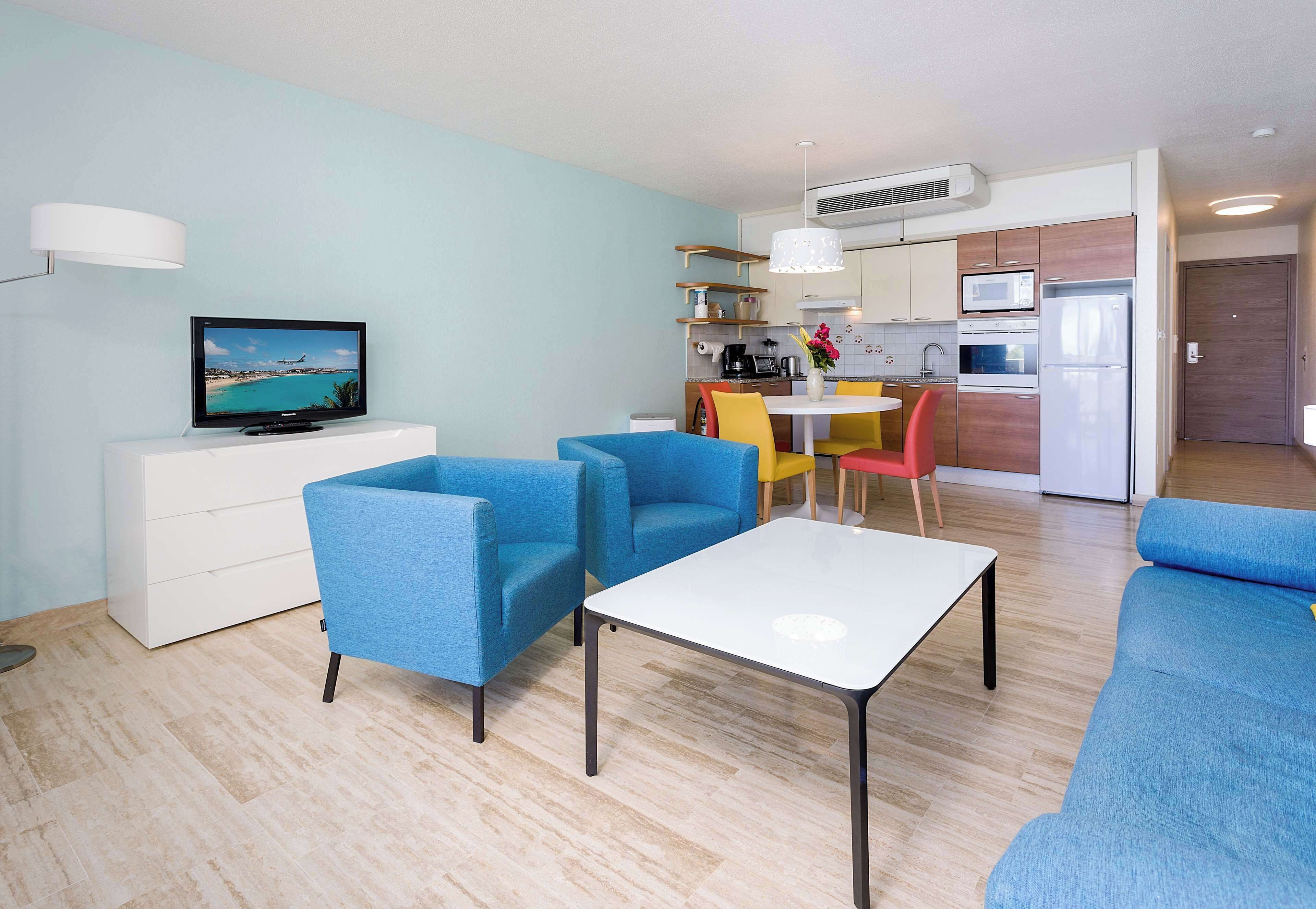 Uma sala de estar moderna com televisão de tela plana, sofás azuis, uma mesa de centro branca e uma área de jantar com uma pequena mesa e cadeiras.