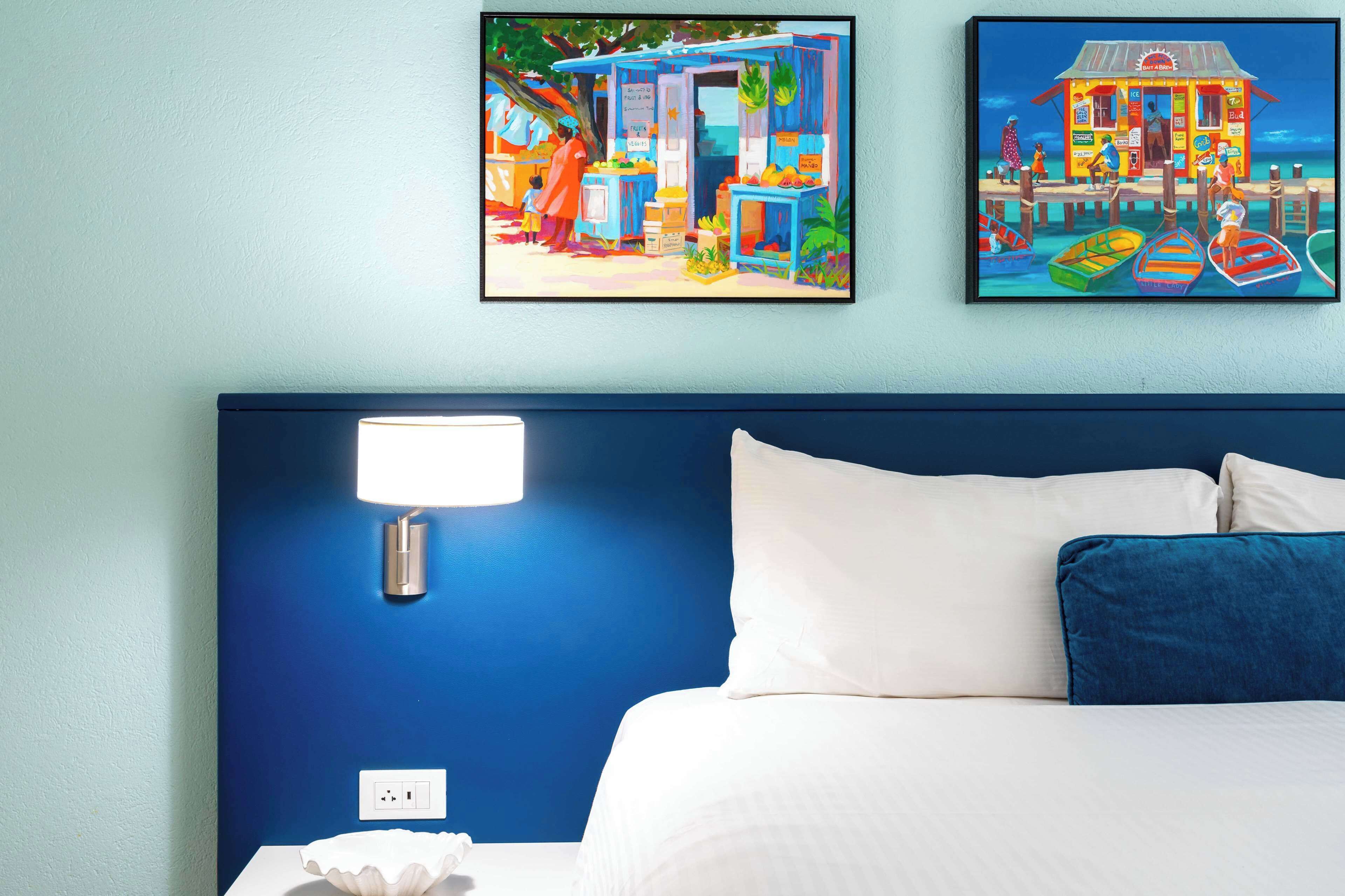 Quarto de hotel com cabeceira azul, roupa de cama branca e duas pinturas retratando paisagens tropicais.