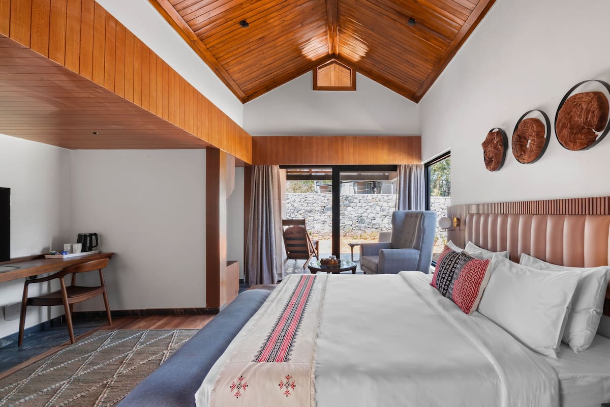 Quarto de hotel moderno com cama grande, teto de madeira e parede de pedra visível pela janela.