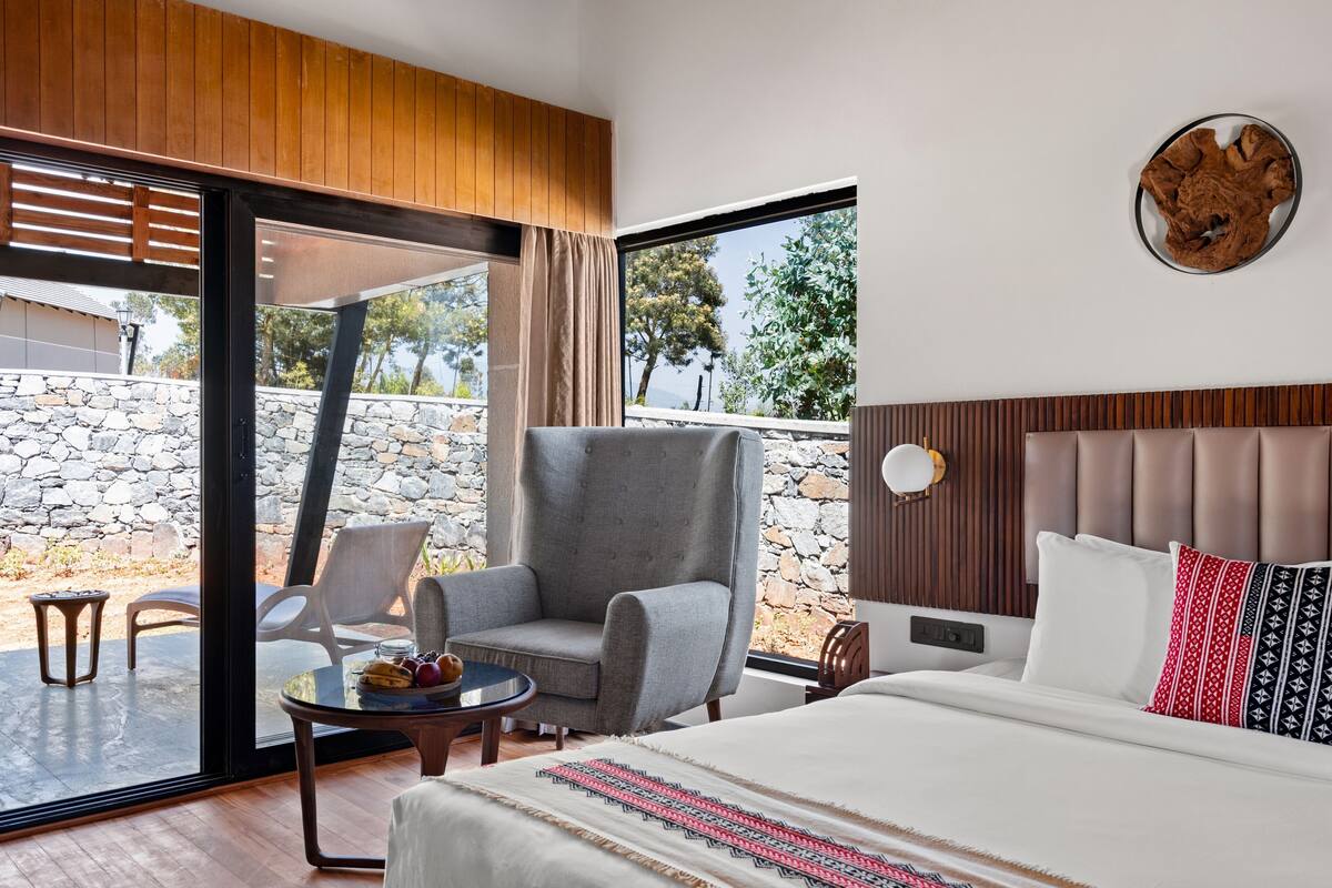 Quarto de hotel moderno com uma cama grande, área de estar com mesa e cadeiras, parede de pedra e vista para o exterior.
