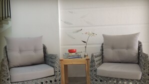 Duas cadeiras de vime cinza com almofadas, uma pequena mesa de madeira com um vaso e livros, e uma parede branca com padrão de folhas.