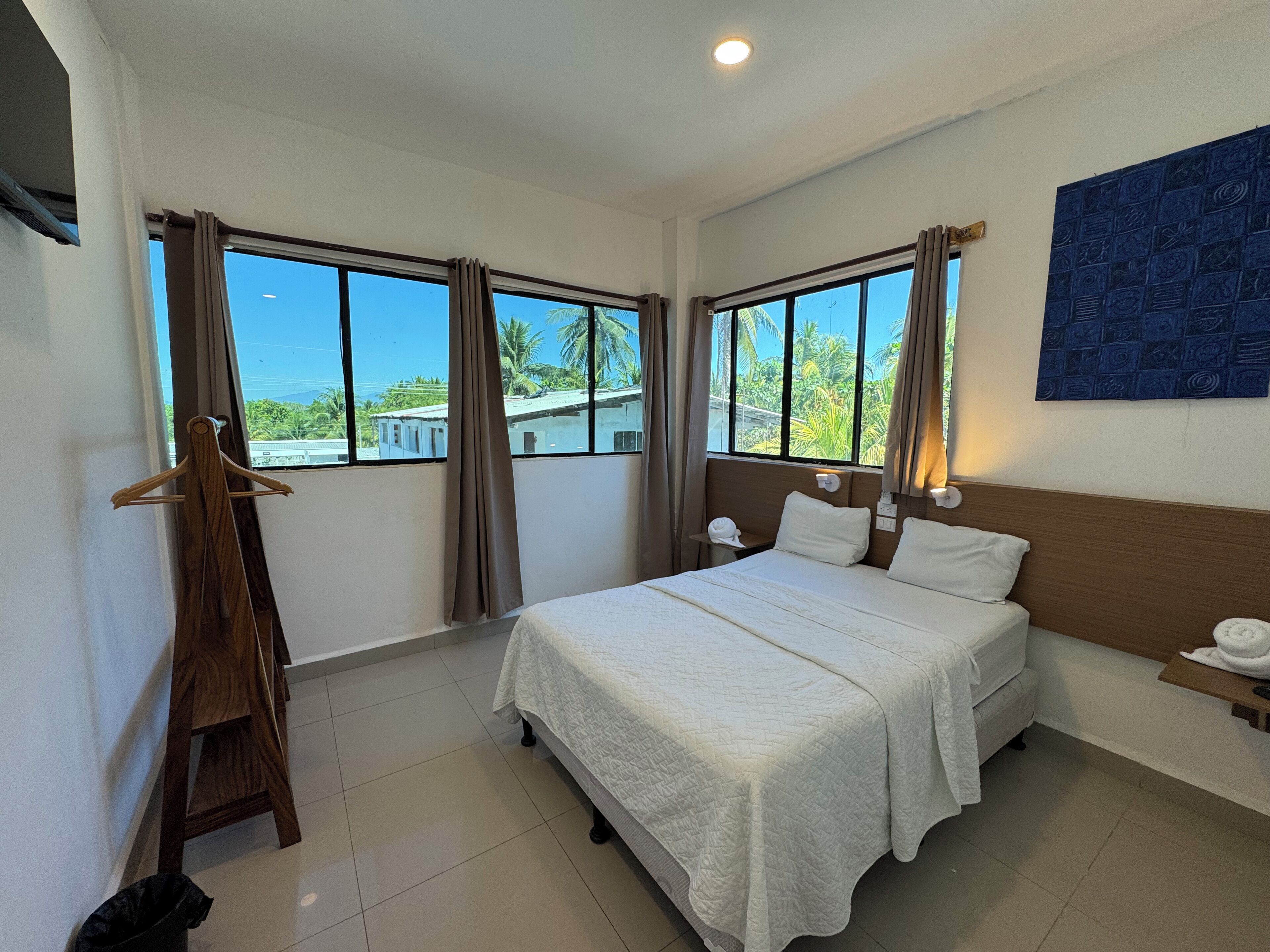 Quarto de hotel com uma cama grande, duas janelas com cortinas, um cabide de madeira, uma mesinha de cabeceira e uma obra de arte na parede.