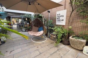 Uma cadeira suspensa sob um guarda-sol, plantas em vasos e uma parede de tijolos com uma placa.