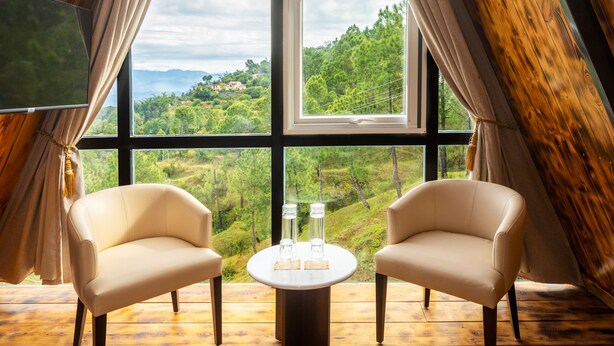 Interior em madeira com duas cadeiras e uma mesa, grandes janelas com vista para áreas verdes.