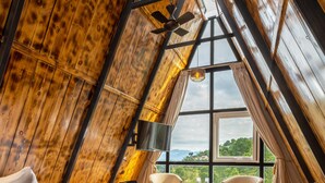 Quarto amplo com pé direito alto, paredes de madeira, uma cama, duas cadeiras, uma pequena mesa e uma grande janela com vista para o exterior.