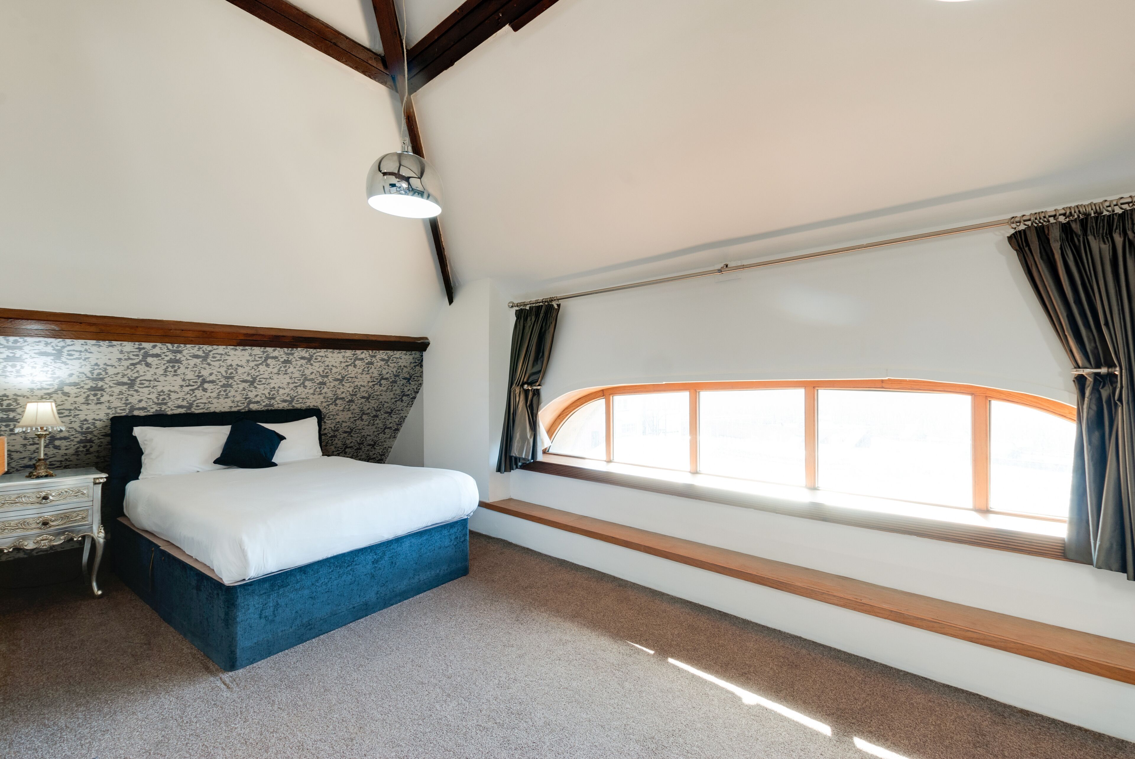 Quarto com cama, mesa de cabeceira, luminária, janela com cortinas e vista para o exterior.