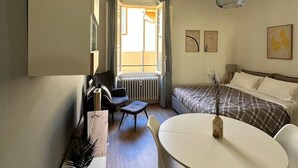 Quarto de hotel pequeno e acolhedor, com cama, mesa de jantar compacta com cadeiras, sofá, radiador e janela com cortinas.