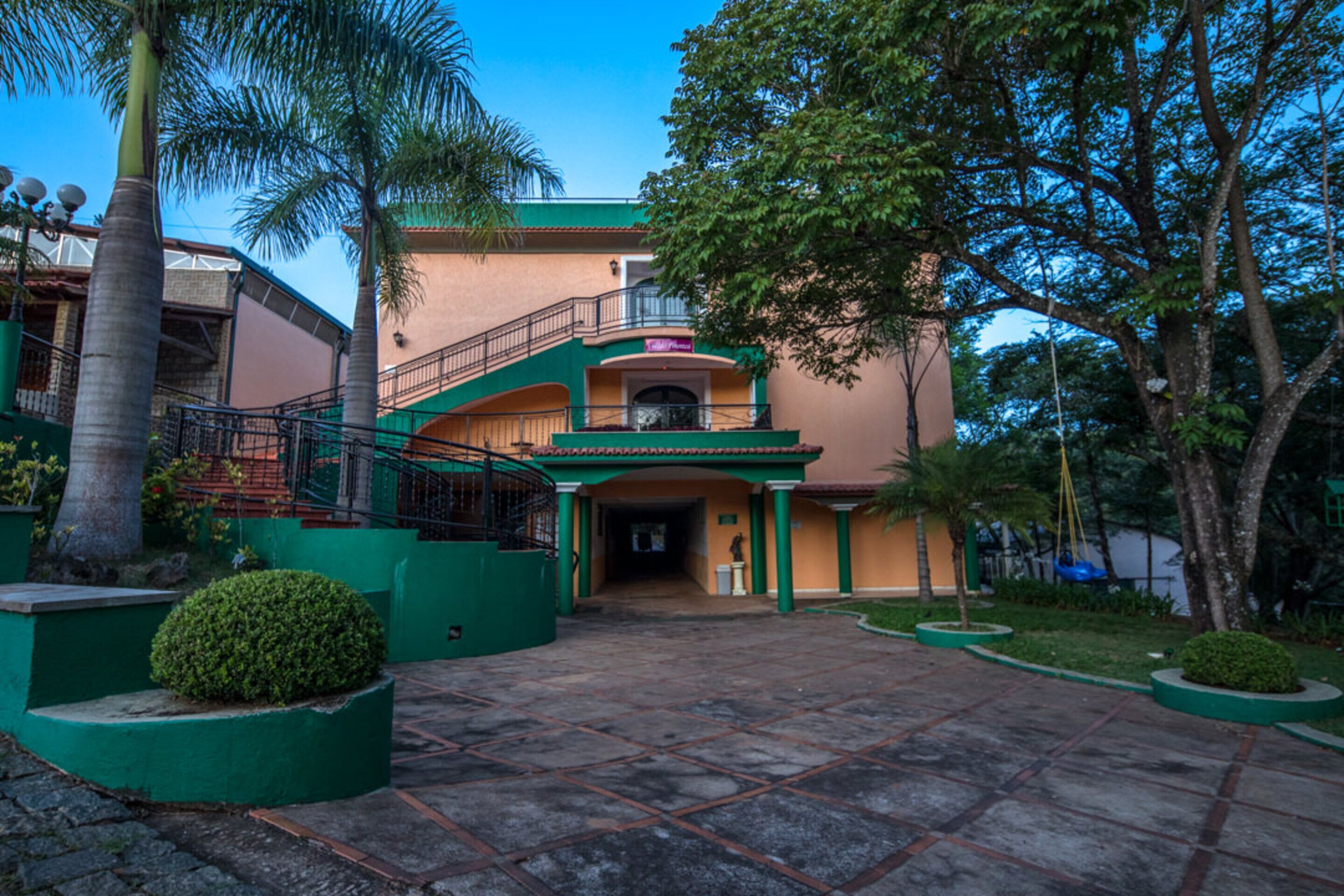 Um edifício com fachada verde e bege, entrada central com escadaria e vegetação ao redor, incluindo palmeiras e arbustos.