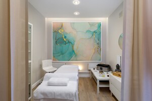 Um spa moderno com uma grande pintura abstrata, uma mesa de massagem, uma cadeira branca e uma escrivaninha com um rádio e uma pequena estátua.