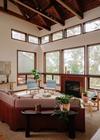 Uma sala de estar espaçosa com um sofá modular grande, duas poltronas, lareira e grandes janelas com vista para árvores.