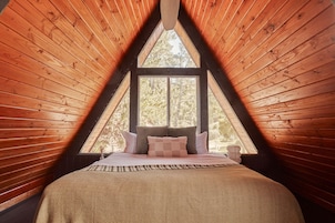 Um quarto de madeira triangular com uma cama, almofadas e uma janela.