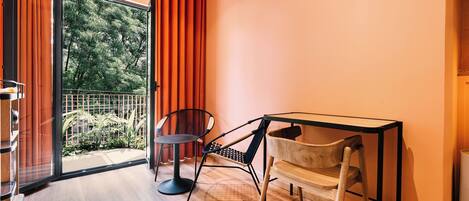 Quarto com piso de madeira, uma mesa de metal preto, uma cadeira de madeira e vista para o verde através de uma porta de vidro.