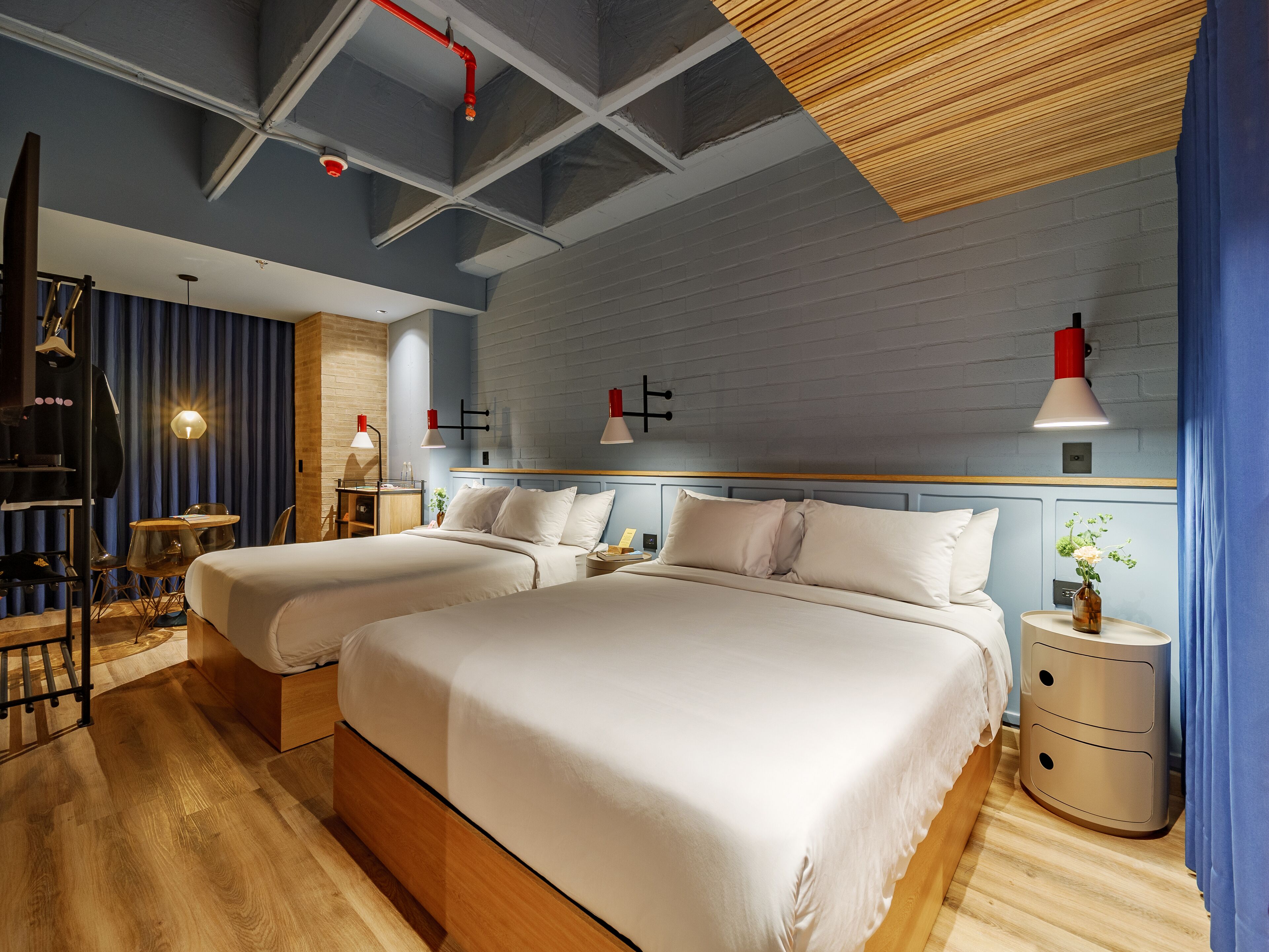 Quarto de hotel moderno com duas camas, piso de madeira, parede de tijolos e cortina azul.