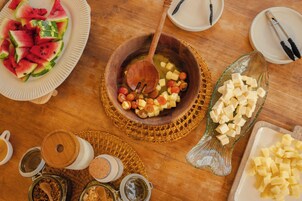 Uma mesa de madeira com diversos alimentos e utensílios, incluindo uma fatia de melancia, uma tigela com uma colher de pau e um recipiente de vidro com cubos de queijo.