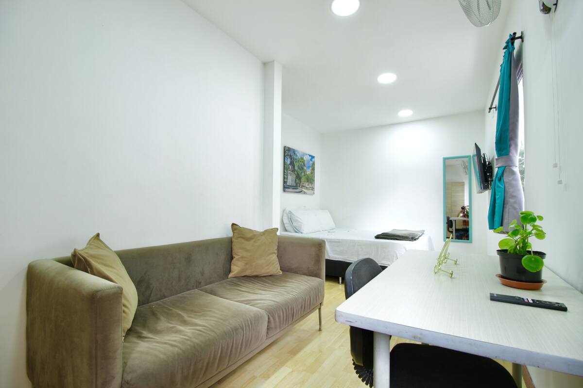 Um quarto moderno e bem iluminado, com um sofá, uma pequena mesa com uma planta e uma cama ao fundo.