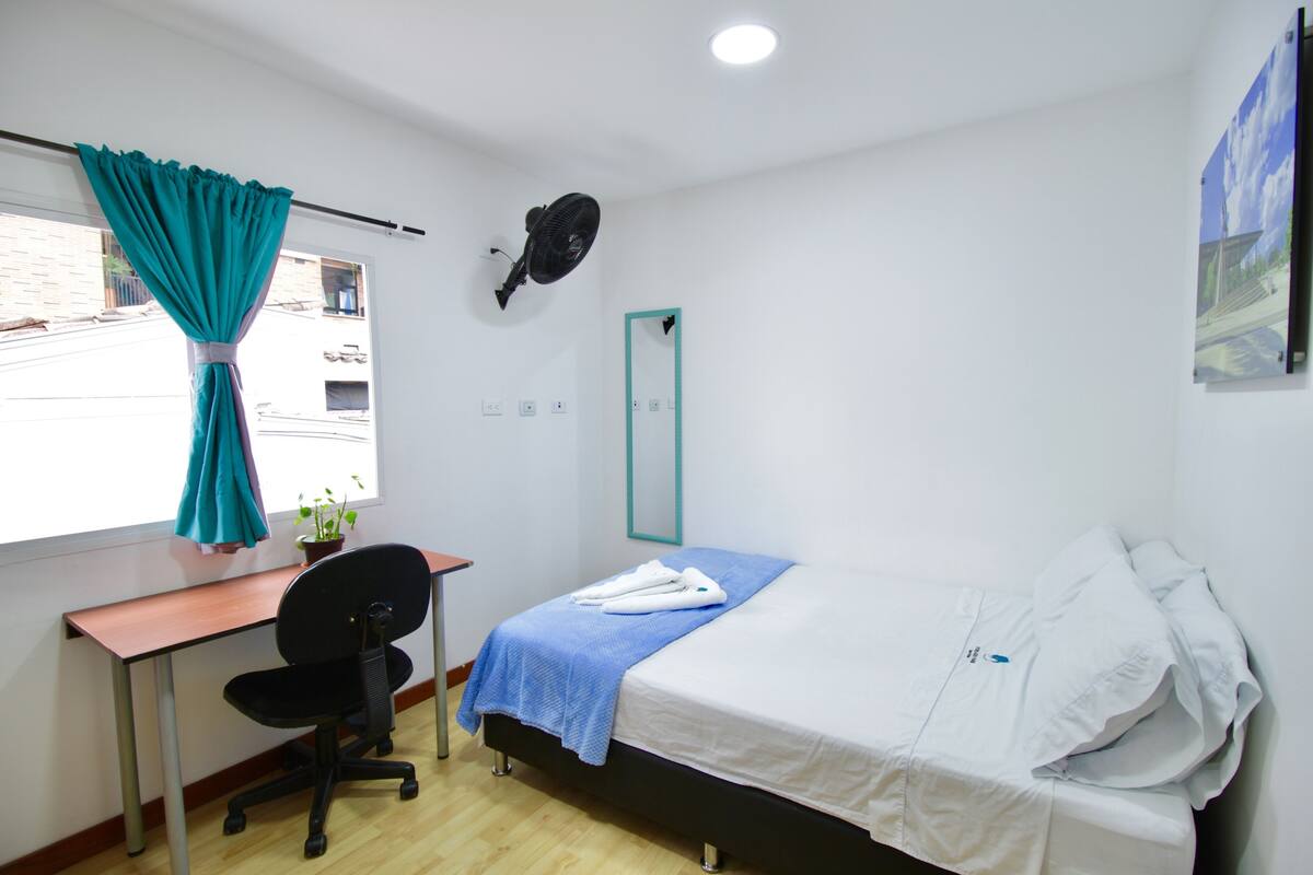 Quarto com cama, escrivaninha, cadeira, planta em vaso, espelho e ventoinha na parede.