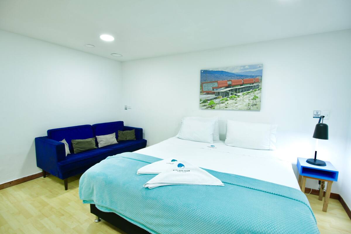Quarto de hotel com cama, sofá azul, mesa de cabeceira com abajur e pintura pendurada na parede.