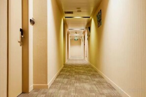 Corredor de hotel com piso revestido de carpete, portas à esquerda e luminária no teto.