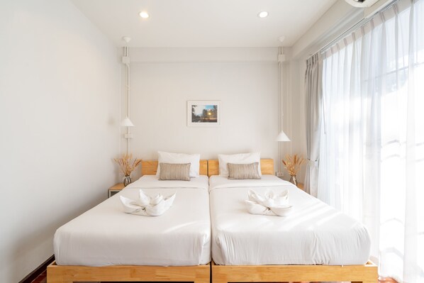 Quarto duplo com duas camas, cada uma com roupa de cama branca e almofadas decorativas, uma pequena moldura na parede e uma janela grande com cortinas translúcidas.