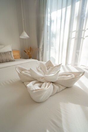 Cama bem arrumada com toalha branca disposta em formato de flor, luminária de cabeceira e janela com cortinas translúcidas.