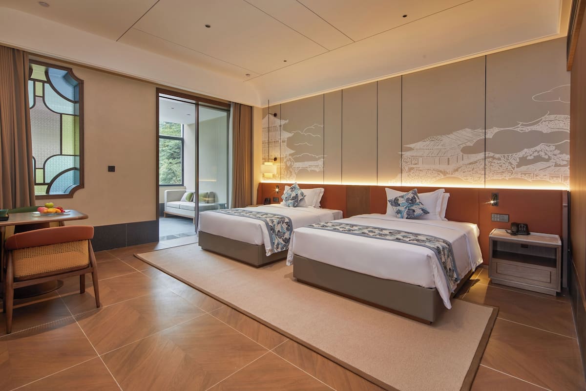 Quarto de hotel com duas camas, uma escrivaninha e vista para o verde através de uma porta de vidro.