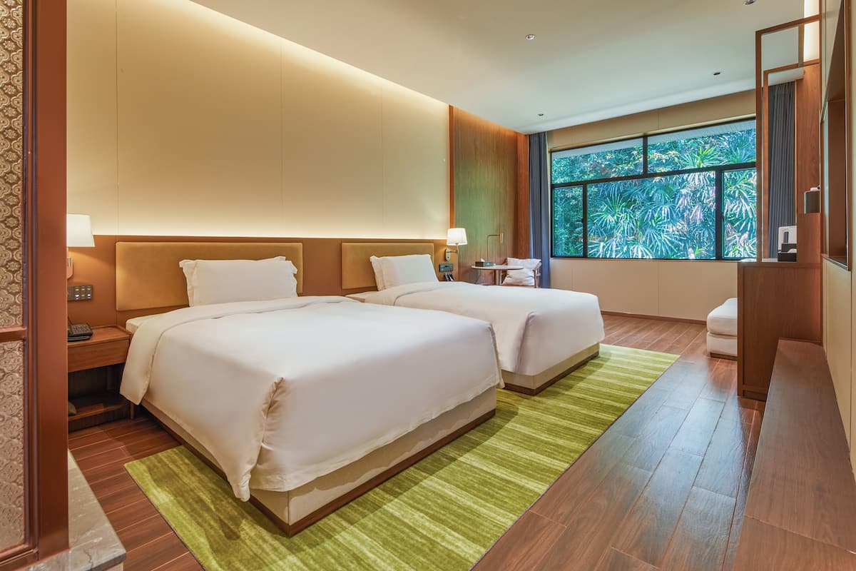 Quarto de hotel com duas camas, uma janela grande com vista para vegetação, tapete verde e piso de madeira.