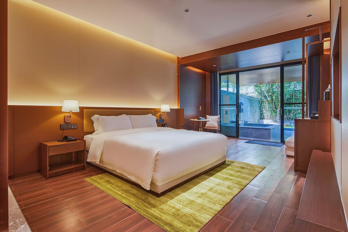 Quarto de hotel moderno com uma cama grande, abajures, uma mesinha e vista para o exterior através de grandes janelas.