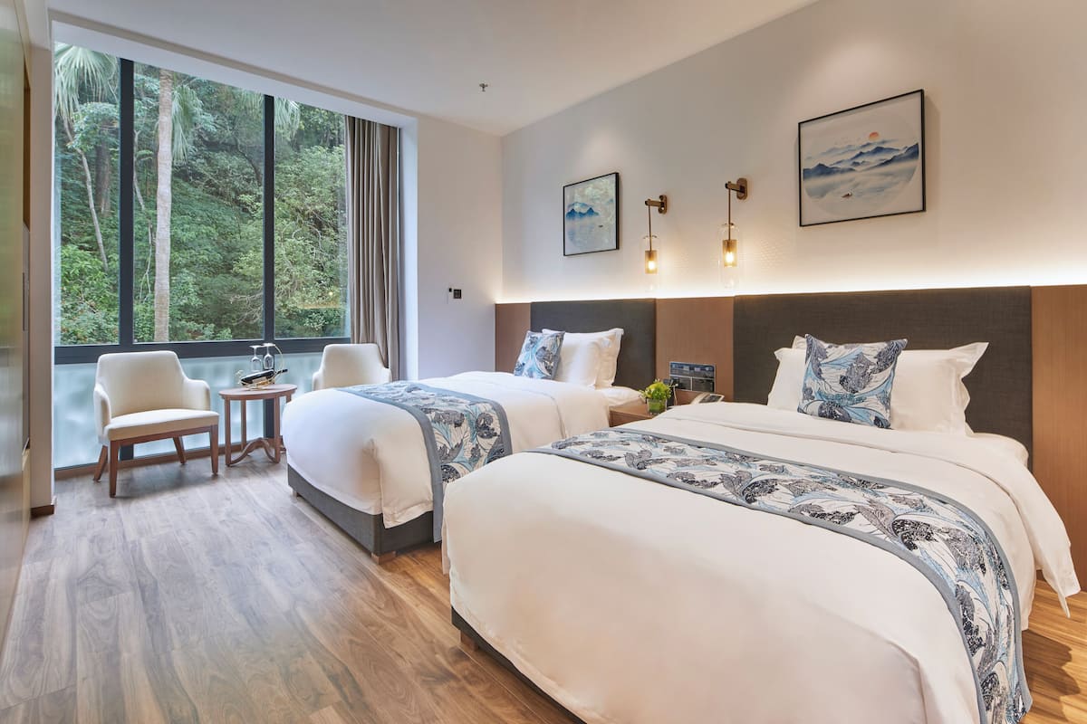 Quarto de hotel com duas camas, área de estar com uma cadeira e mesa, e amplas janelas com vista para a floresta.