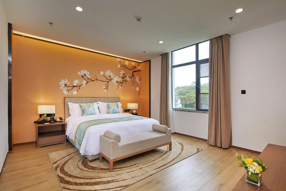 Quarto de hotel moderno com uma cama grande, mesas de cabeceira, uma parede decorativa, uma janela ampla com cortinas e uma pequena mesa com flores.