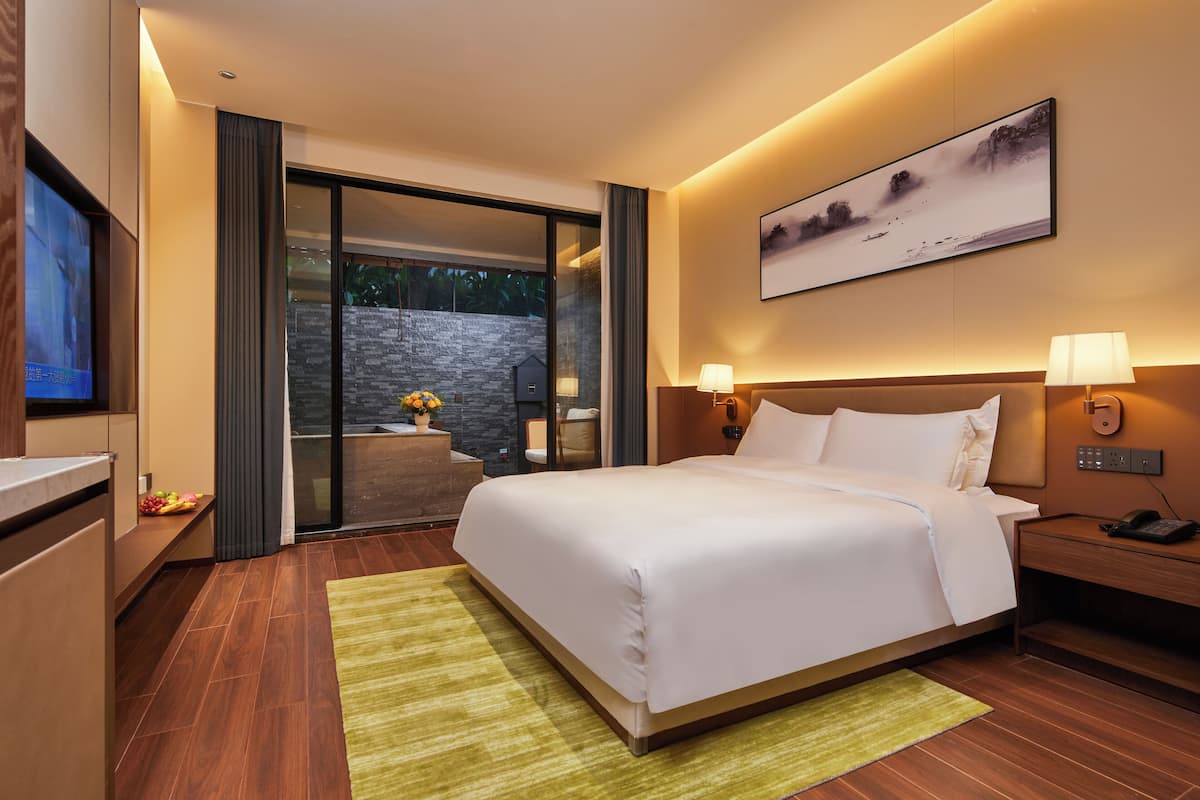 Quarto de hotel moderno com uma cama grande, televisão, escrivaninha com abajur e vista para o exterior por uma porta de correr.