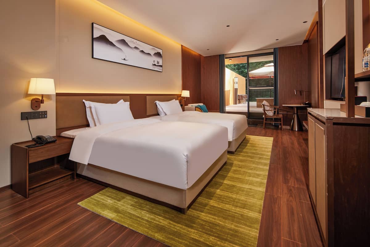 Quarto de hotel com duas camas, uma escrivaninha, uma cadeira, uma televisão e uma janela grande.