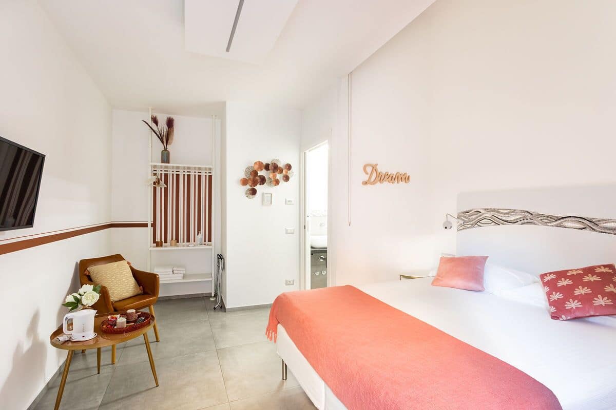 Quarto de hotel com cama, uma poltrona, uma mesinha com chaleira e flores, decoração na parede e uma placa com a palavra 'Dreams'.