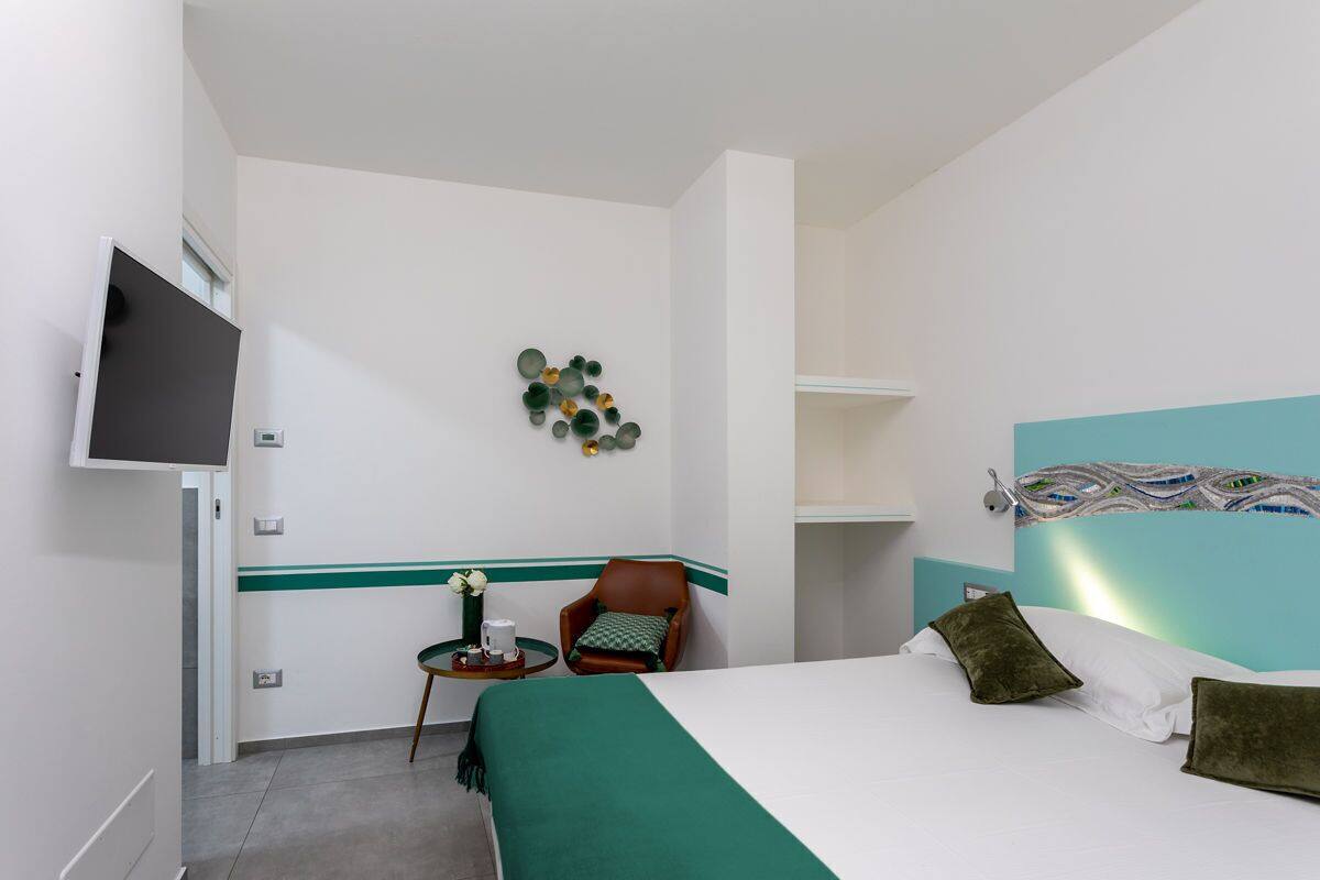 Quarto de hotel com cama, televisão, uma mesinha com uma chávena, cadeira e um objeto decorativo na parede.