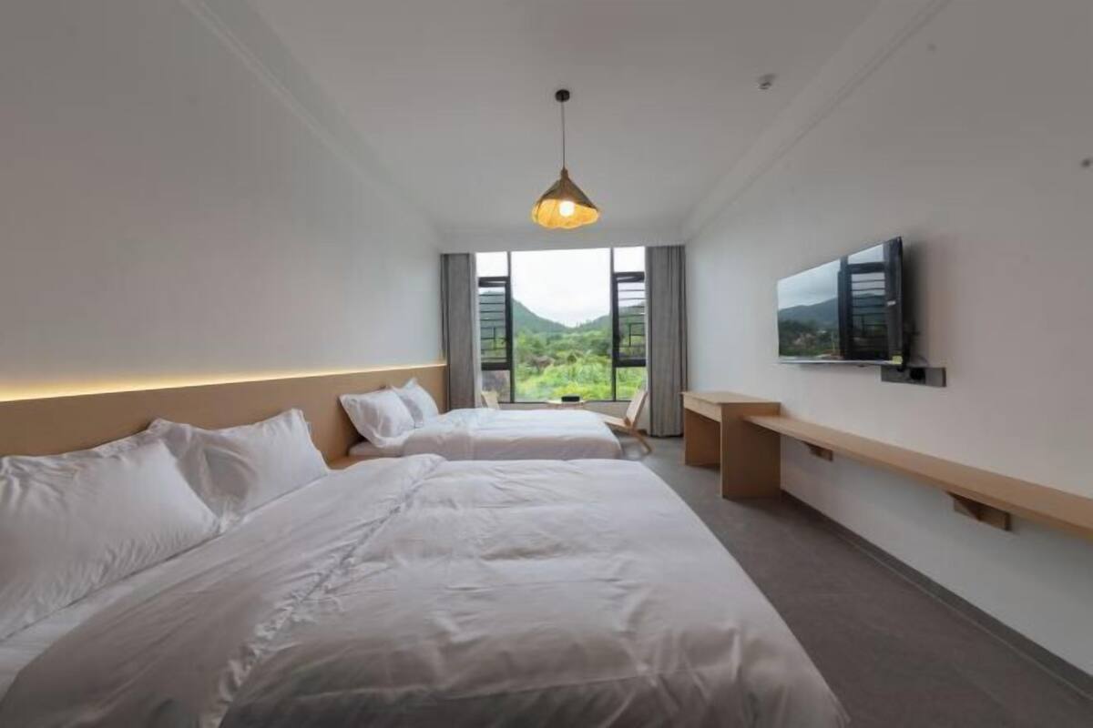 Quarto de hotel com duas camas, televisão, janela com vista e uma luminária pendente.
