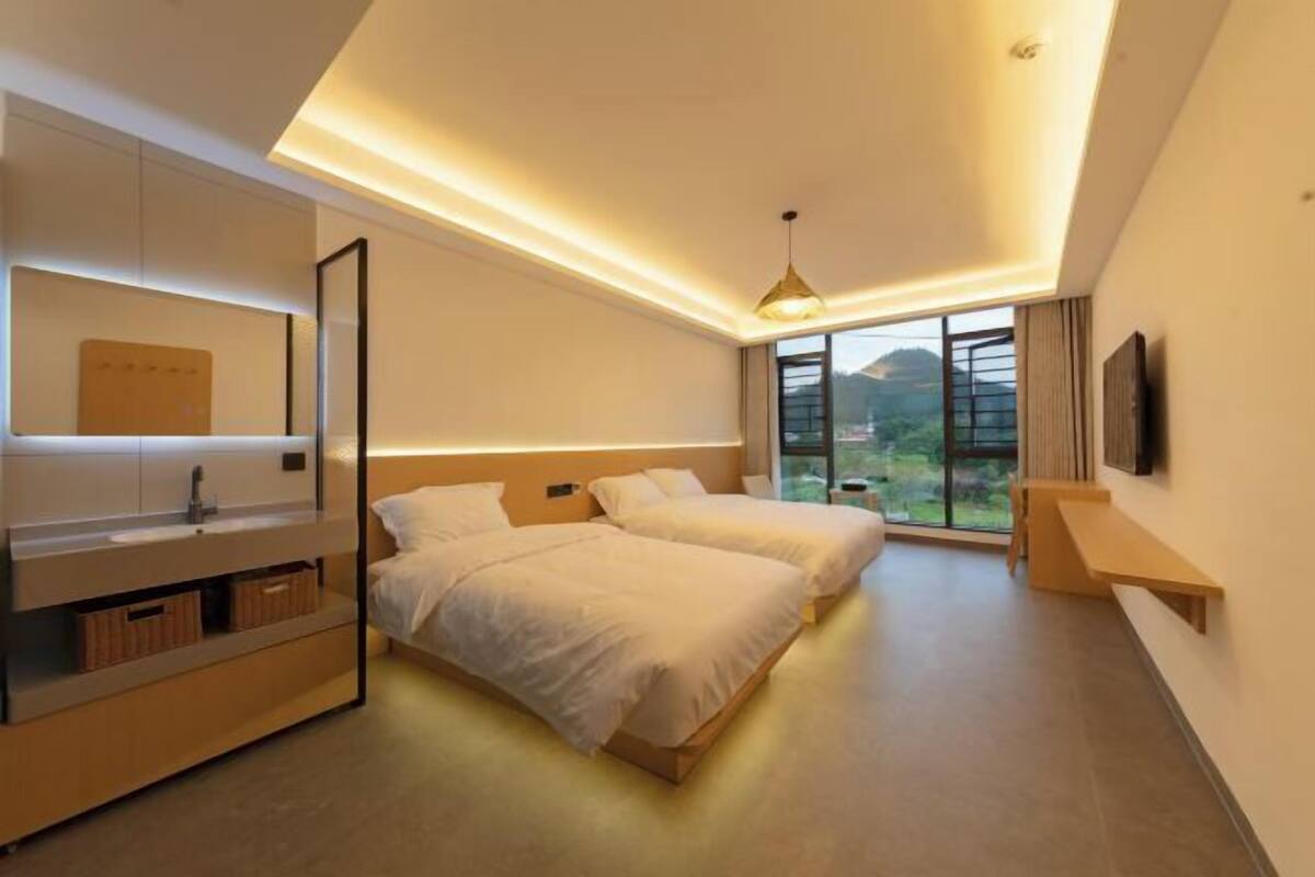 Quarto moderno com duas camas, guarda-roupa embutido, uma janela ampla com vista e uma luminária pendente.