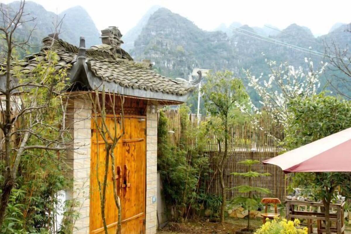 Um edifício tradicional com telhado em cerâmica e porta de madeira, rodeado por vegetação exuberante e montanhas ao fundo.