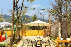 Um pátio externo com mesa e cadeiras sob um guarda-sol grande, cercado por árvores e um muro de madeira.