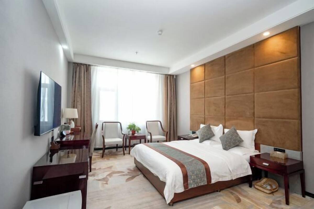 Quarto de hotel moderno com uma cama grande, escrivaninha, cadeira, televisão e janela com cortinas.