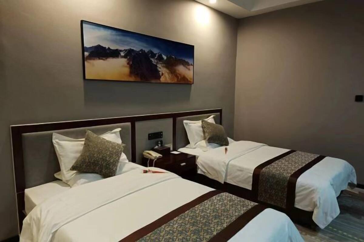 Quarto de hotel com duas camas, uma mesa de cabeceira, um telefone e um quadro de montanhas na parede.