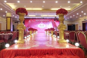 Salão de banquetes com mesas dispostas em fileiras, palco com fundo rosa e pilares dourados adornados com flores vermelhas.