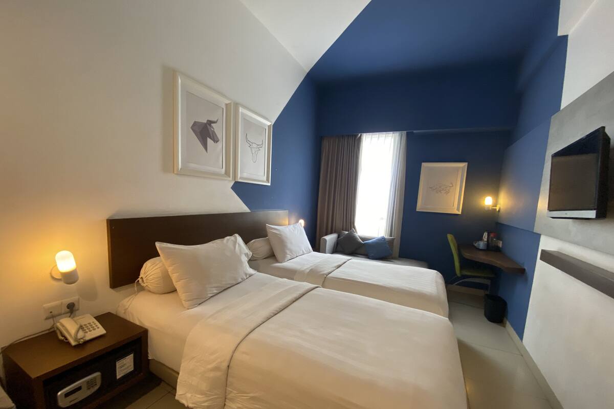 Quarto de hotel com duas camas, uma mesa de cabeceira com telefone, uma escrivaninha pequena com cadeira, televisão e obras de arte nas paredes.
