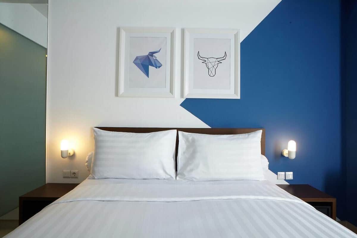 Quarto de hotel com uma cama, duas mesas de cabeceira e duas luminárias fixadas na parede.