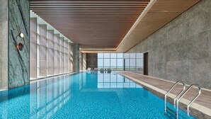 Piscina interior moderna com água azul cristalina, deck de madeira e vista para o horizonte urbano através de grandes janelas.