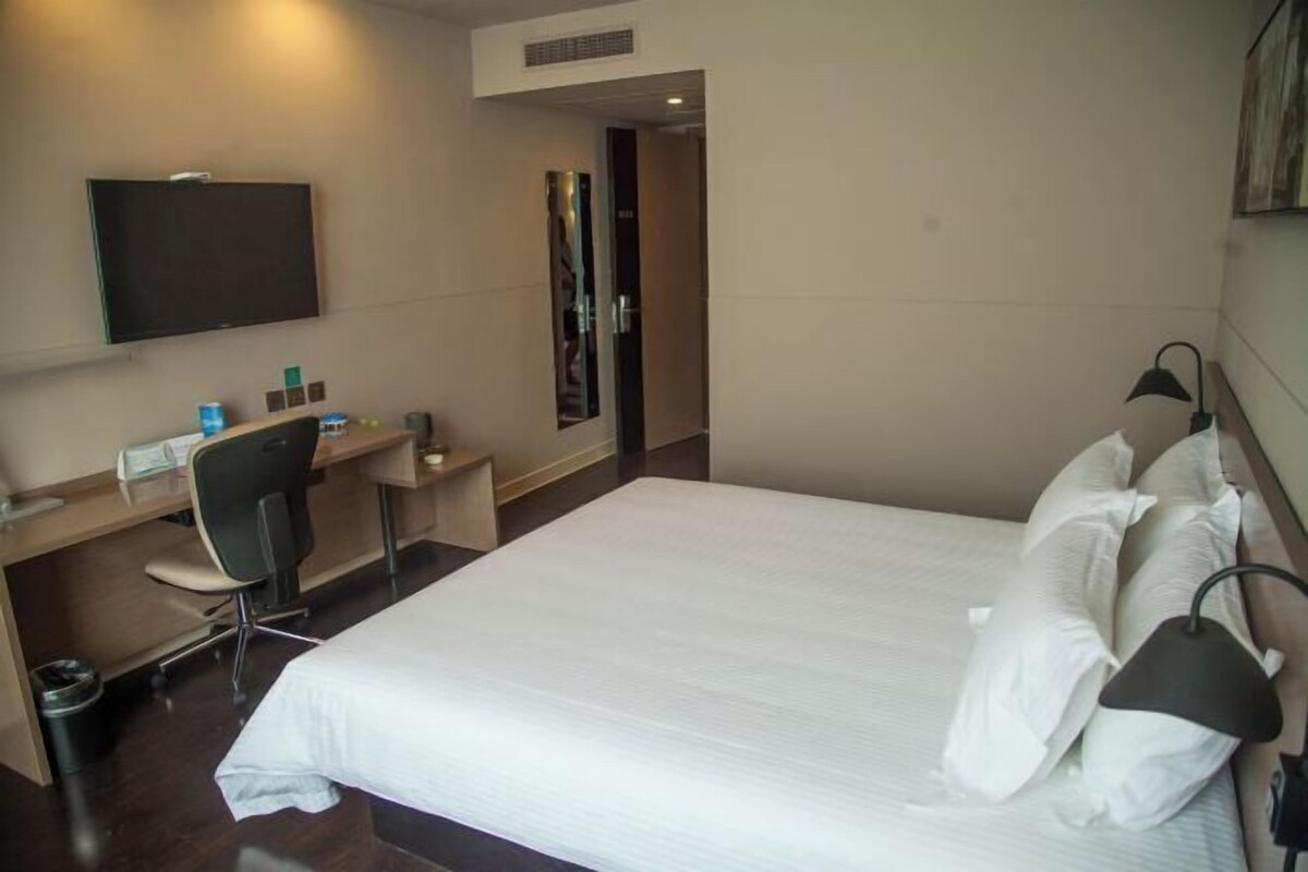 Quarto de hotel com cama, escrivaninha, cadeira, televisão e espelho.