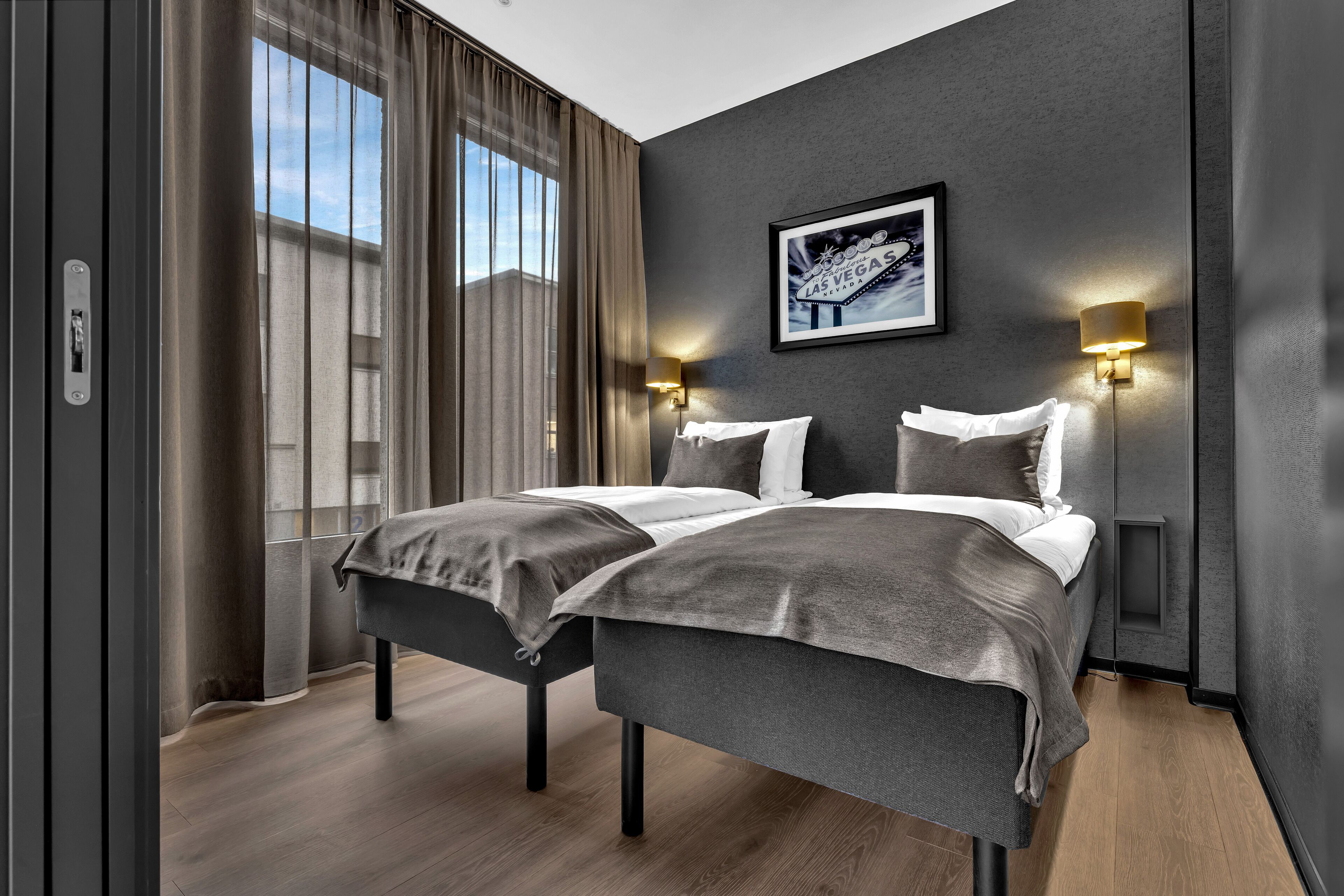Quarto de hotel moderno com duas camas, uma janela grande com cortinas, um quadro com a placa de Las Vegas e duas luminárias fixadas na parede.