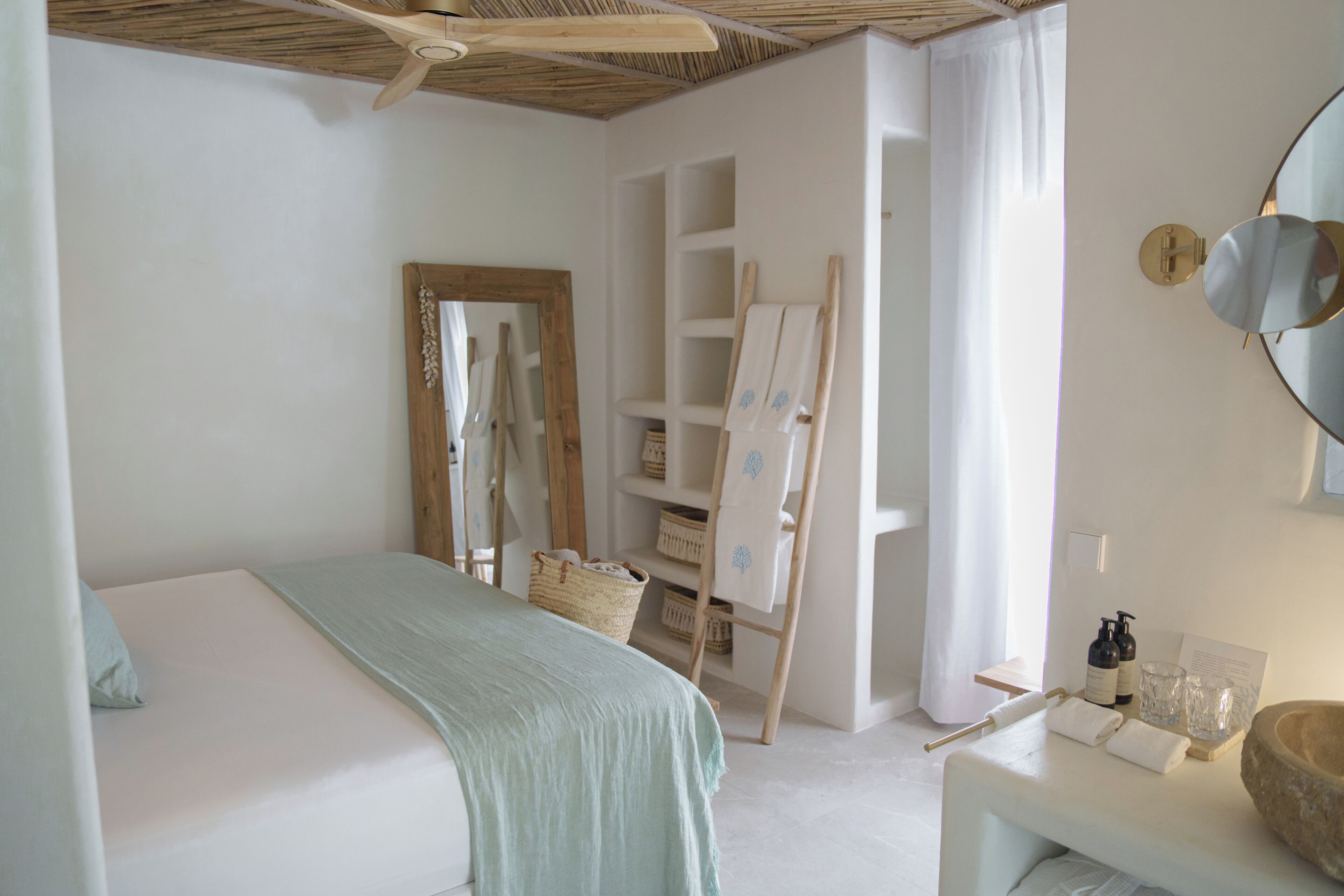 Quarto com cama, ventoinha de teto em madeira, espelho, prateleira com toalhas e pequena mesa com artigos de higiene.