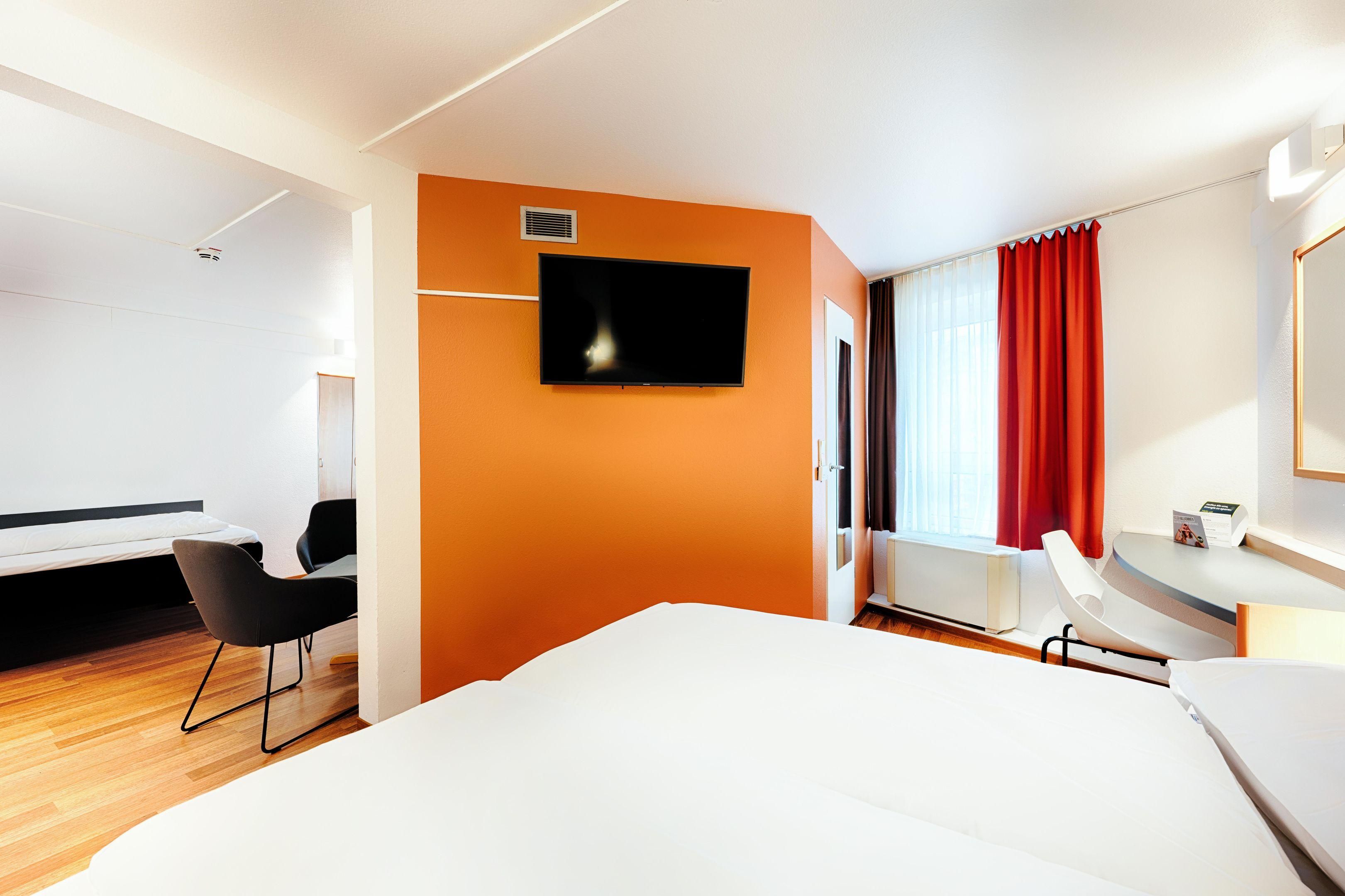 Quarto de hotel com uma cama, uma escrivaninha, uma cadeira, uma televisão e uma janela com cortinas.