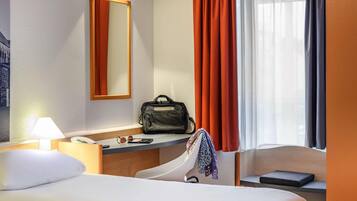 Quarto de hotel com cama, escrivaninha com luminária, cadeira, mala e janela com cortinas.