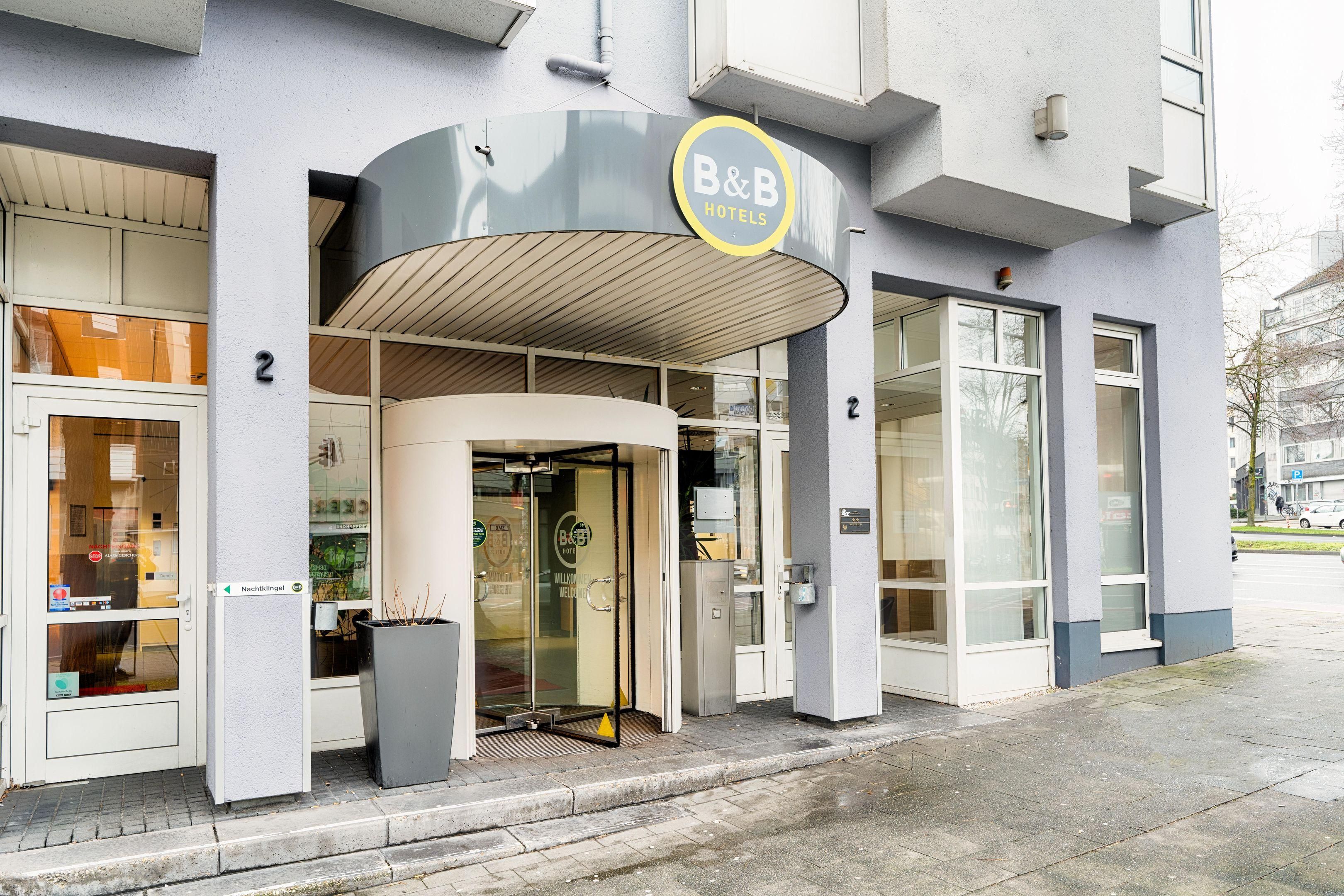 Entrada de hotel com marquise curva, portas de vidro e placa indicando "B&B Hotels".