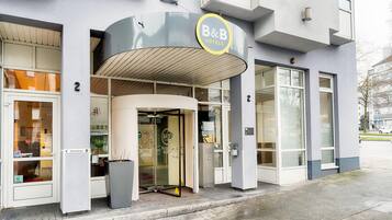 Entrada de hotel com marquise curva, portas de vidro e placa indicando "B&B Hotels".